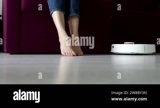 robot vacuum cleaner driving around barefoot woman at home. Female lift ...