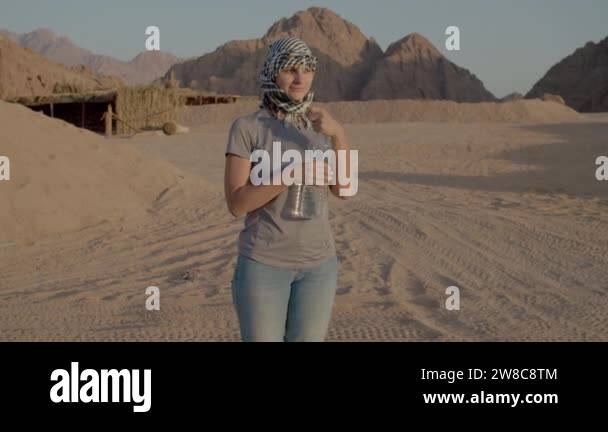 Woman in checkered keffiyeh drinking water of plastic bottle standing ...