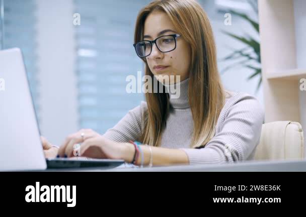 Cute woman working at a laptop with glasses, typing text in the office ...