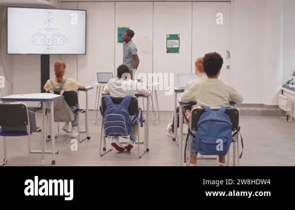 Wide shot of young Black male teacher standing in classroom, explaining ...