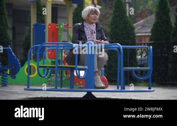 Wide shot thoughtful sad senior beautiful woman sitting on merry-go-round on playground outdoors ...