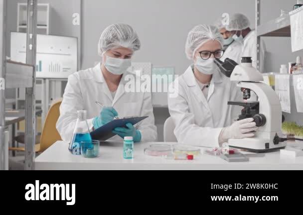 Waist-up of two female scientists wearing face masks, gloves, medical gowns and hats sitting at ...