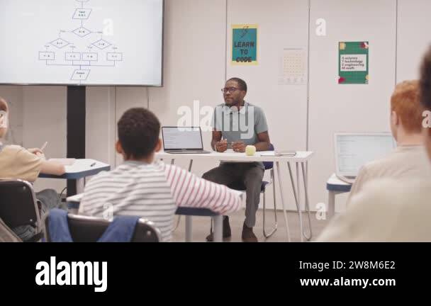 Wide shot of young Black male teacher sitting at desk in classroom ...