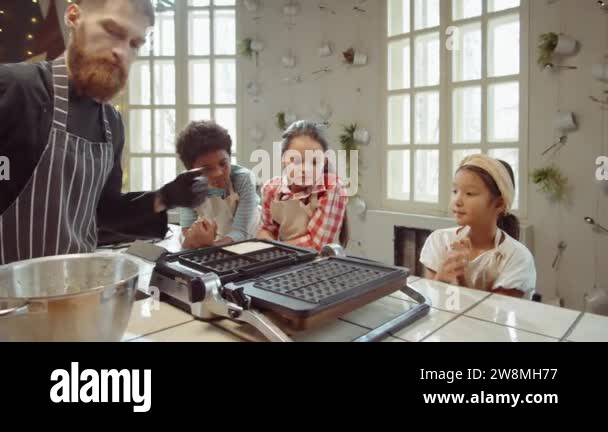 Little girl pouring batter in waffle maker while chef and other kids ...