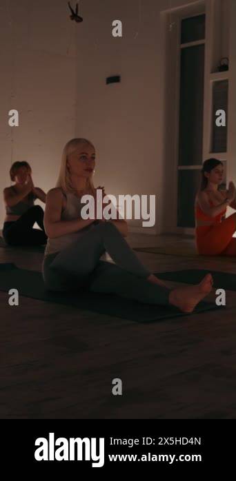 Vertical screen: Individuals in dimly lit yoga studio practice seated ...