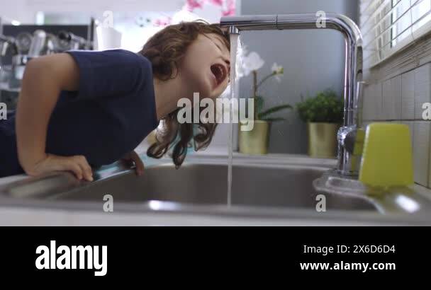 Cute boy with curly hair leans over a kitchen sink to drink water ...