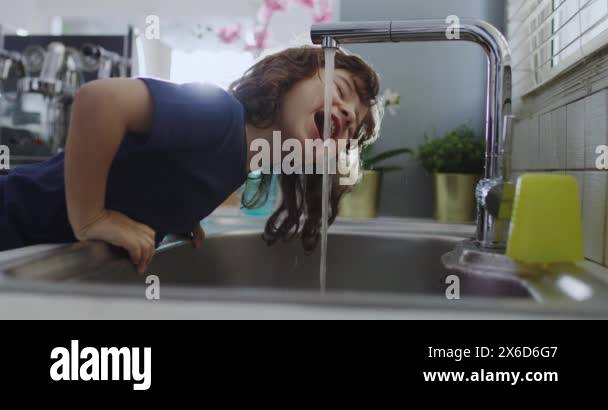 Cute boy with curly hair leans over a kitchen sink to drink water ...