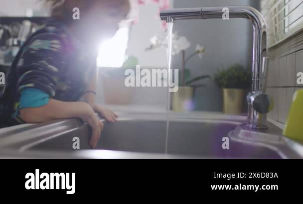 Cute boy with curly hair leans over a kitchen sink to drink water ...