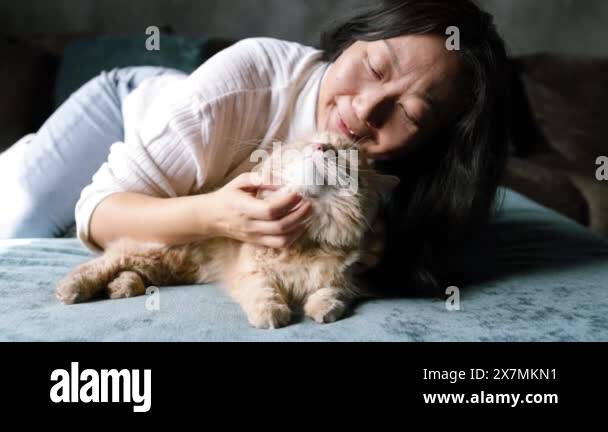 Woman petting cat on sofa in living room. Asian female owner stroking ...