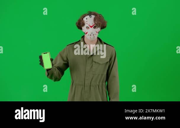 Maniac in hockey mask and jumpsuit holds smartphone with green screen ...