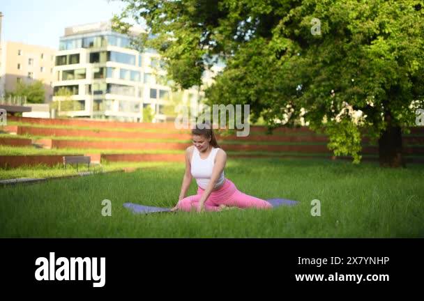 A woman is seated on a yoga mat outdoors in a park. She is engaged in ...