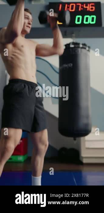 A fit man in a gym concentrated on lifting dumbbells with a punching ...