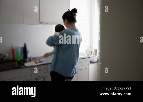Mother multi-tasking in the kitchen, holding her baby while preparing a ...