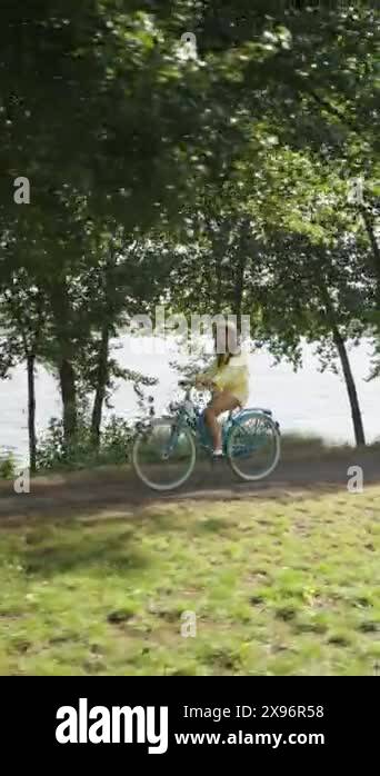 Vertical: Woman rides bicycle along riverside or lakeside path, wearing ...