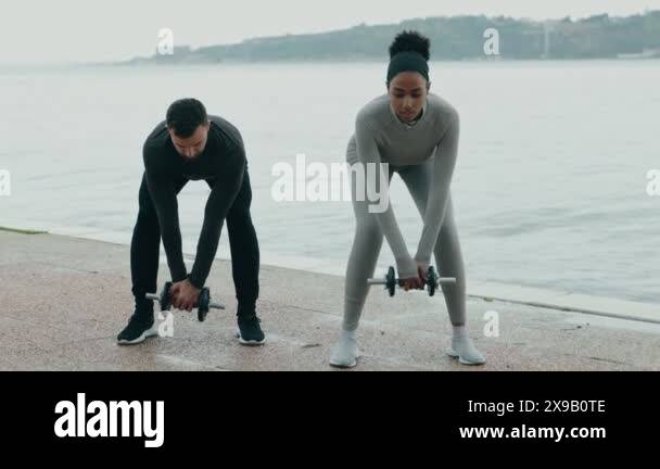 A diverse couple in athletic wear lifts weights together by the riverside training back muscles ...