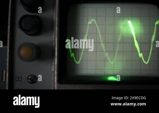 A hand manipulates knobs on a vintage oscilloscope, adjusting the green ...