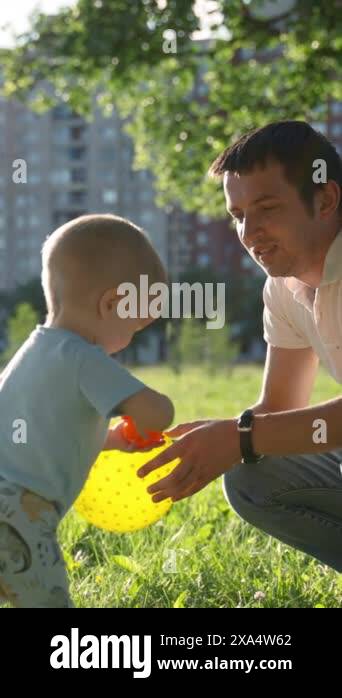 Toddler son and father play with bright balls in green city park ...