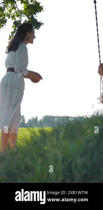 Two little girls swing on a swing suspended from an oak branch. Mom in ...