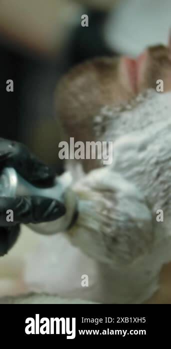 Crop barber in gloves using brush to smear foam on cheek and neck of ...