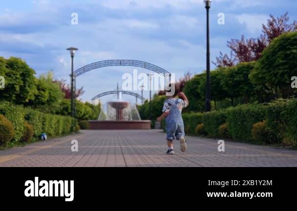 Baby boy runs to the ball lying on the paved road. Green trees, bushes ...