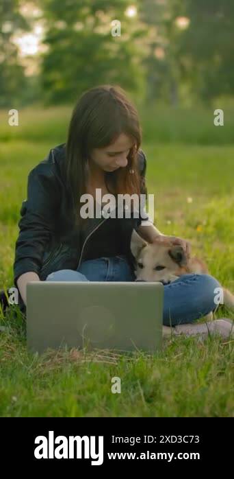 A girl in a park next to an open laptop is petting her dog. Consuming ...