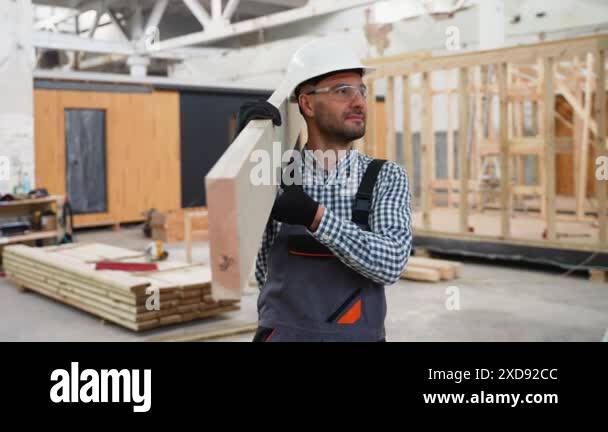 Young carpenter carries a plank across an unfinished prefabricated ...