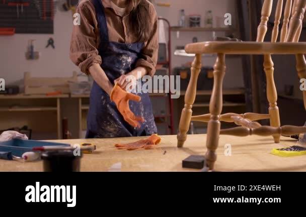 Young woman in atmospheric workshop preparing to restore an old wooden chair, putting on work ...