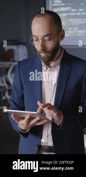Medium vertical portrait of young Middle Eastern man in glasses, jacket using tablet computer ...