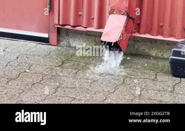 A red metal gutter overflowing with rainwater cascades onto a brick ...