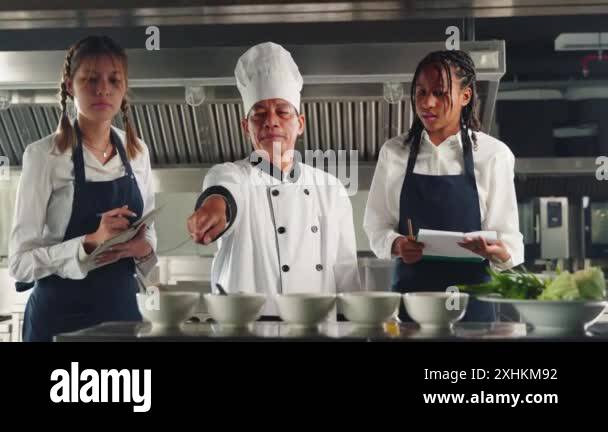 Senior Asian chef in a restaurant kitchen instructs multiracial ...