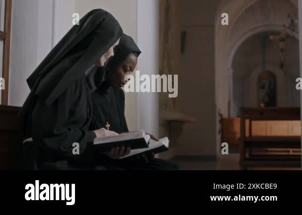 Side view of bi-ethnic sisters in black robes praying on holy bibles ...