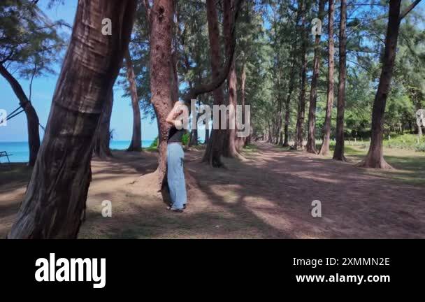 Slow motion video of a young woman walking under the shade of a pine tree by the sea. Tunnel of ...