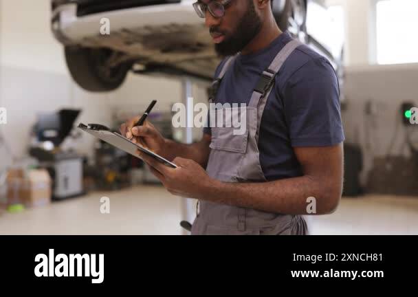 Mechanic carefully inspecting car in auto repair shop with clipboard. Focused professional ...