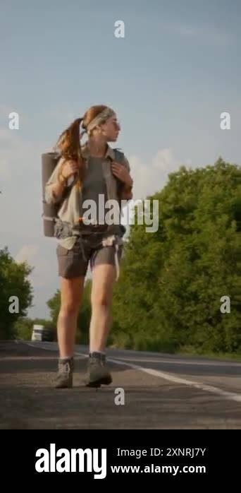 Full vertical shot of young Caucasian female hitchhiker with backpack walking on roadside among ...