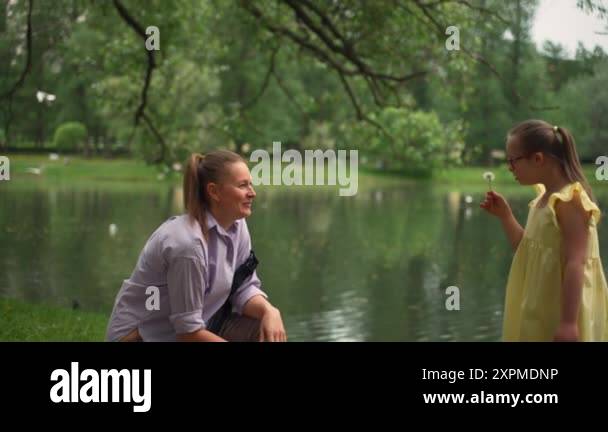 Young girl with Down syndrome in yellow dress is blowing dandelion seeds by lake while mother ...