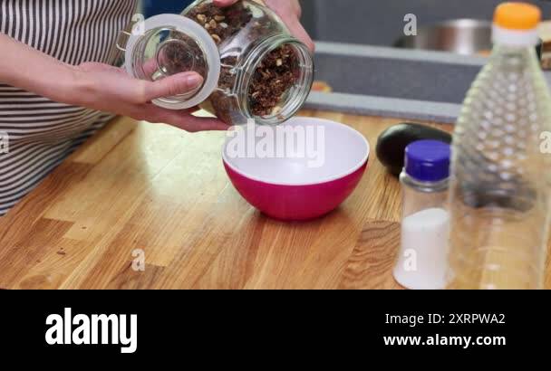 Woman hands hold glass jar pouring mix of nuts into bowl at countertop ...