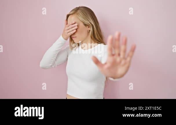 Terrified young blonde woman, covering eyes with hand, making stop ...