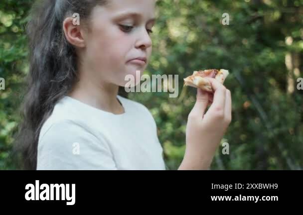 Close up of cute preteen girl eating pizza outdoors in summer. Caucasian child eating fast food ...
