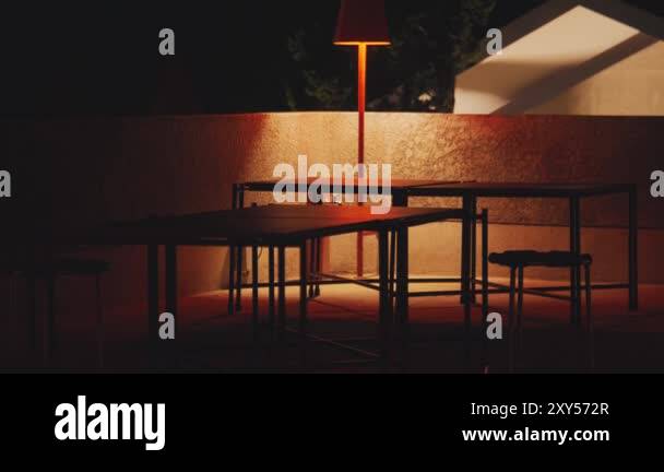 A dimly lit outdoor space featuring several tables and a lamp. The warm ...
