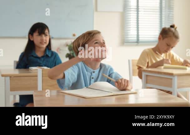 Caucasian smart boy raising hand for asking and answering teacher surrounded by diverse children ...