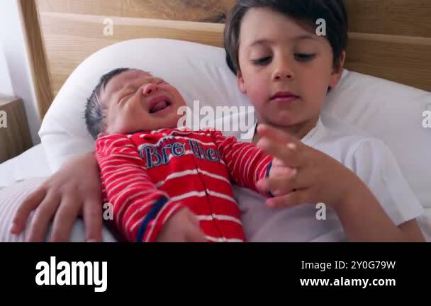 Older brother holding his crying newborn sibling on the bed, showing a ...