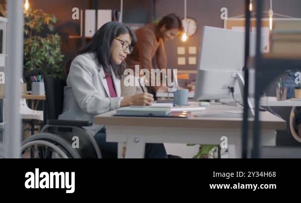 Disabled young lady in wheelchair working in office looking at computer screen and writing in ...