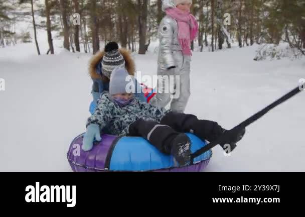 Tracking shot of two boys pulling little girl up hill in snow tube to ...