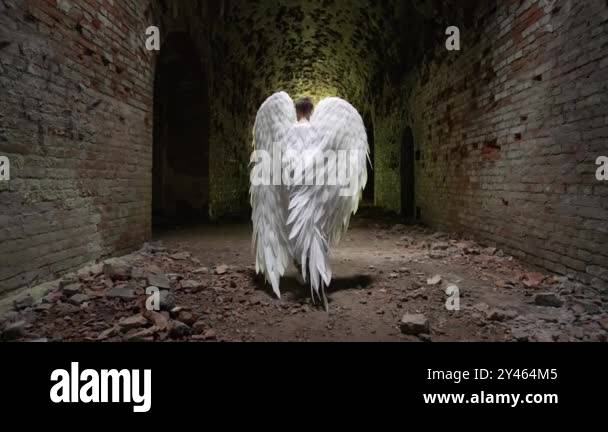 Man in snow-white angel wings stands with his back in abandoned room ...