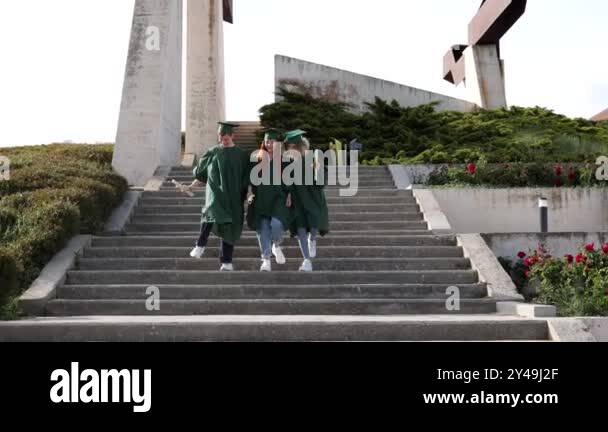 Three young adults in green graduation gowns walk down concrete stairs ...