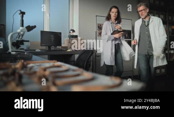 Two archaeologists work in advanced archaeological lab. Mature scientist shows fossil bones of ...