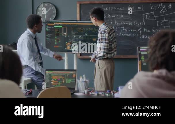 Medium shot of Black male programming teacher and teenage Asian student looking at monitor ...