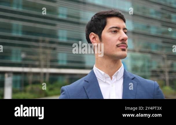 A businessman in a blue suit smiling confidently in front of a modern glass office building. The ...