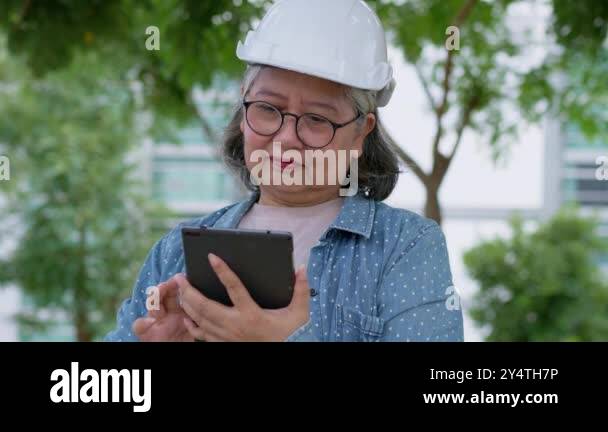 Positive age Asian female architecture in hardhat hold tablet and standing in front of city for ...