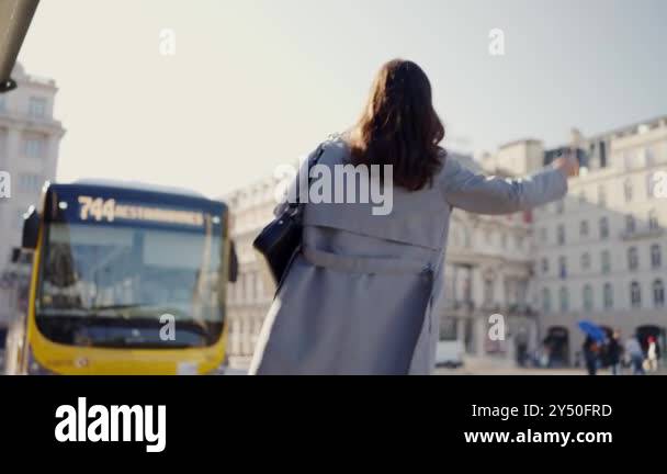 A young woman, viewed from behind, waves for a bus in a vibrant city ...